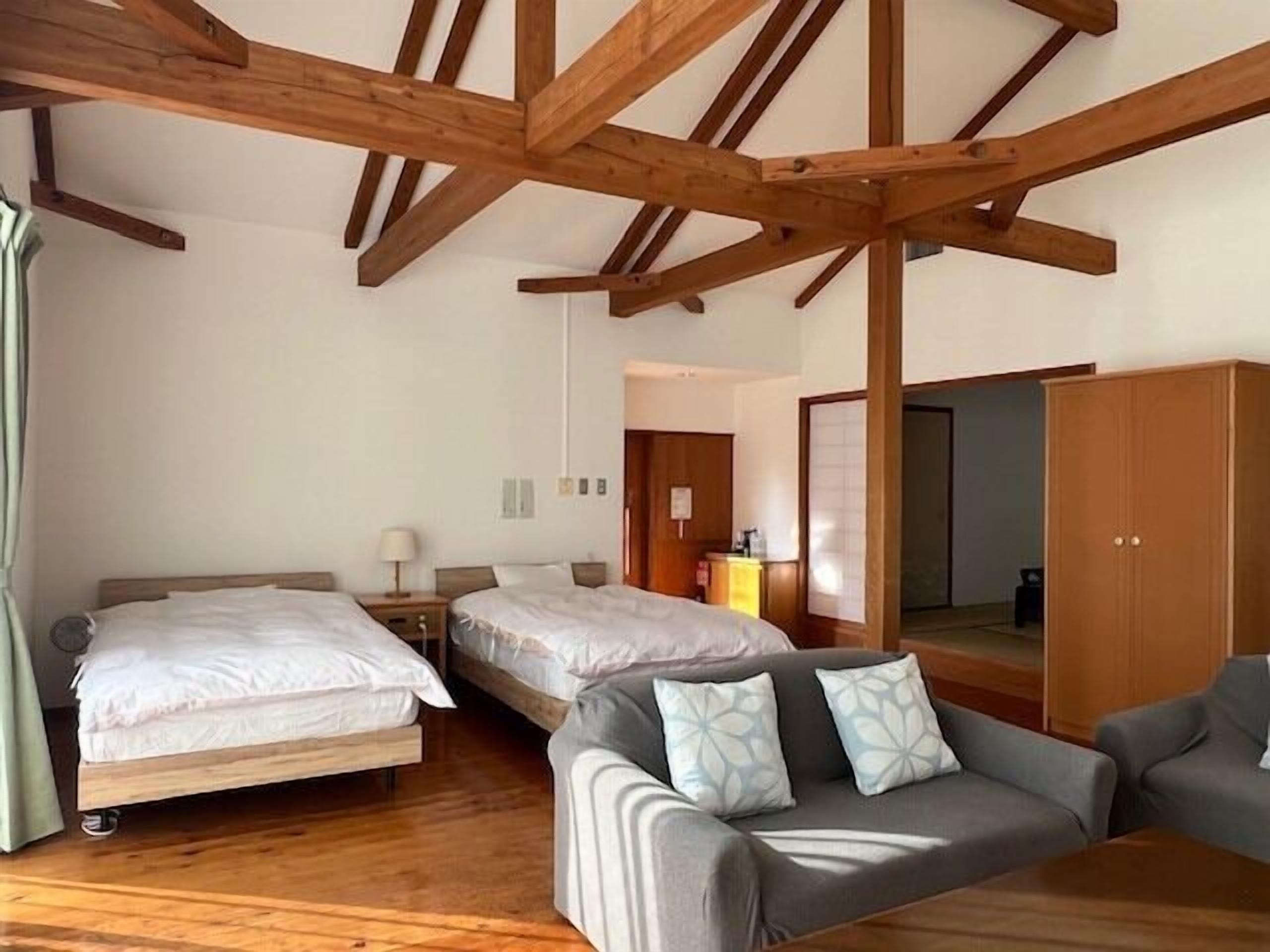 Quarto com duas camas, um sofá, vigas de madeira no teto, um guarda-roupa e uma pequena mesa com luminária.