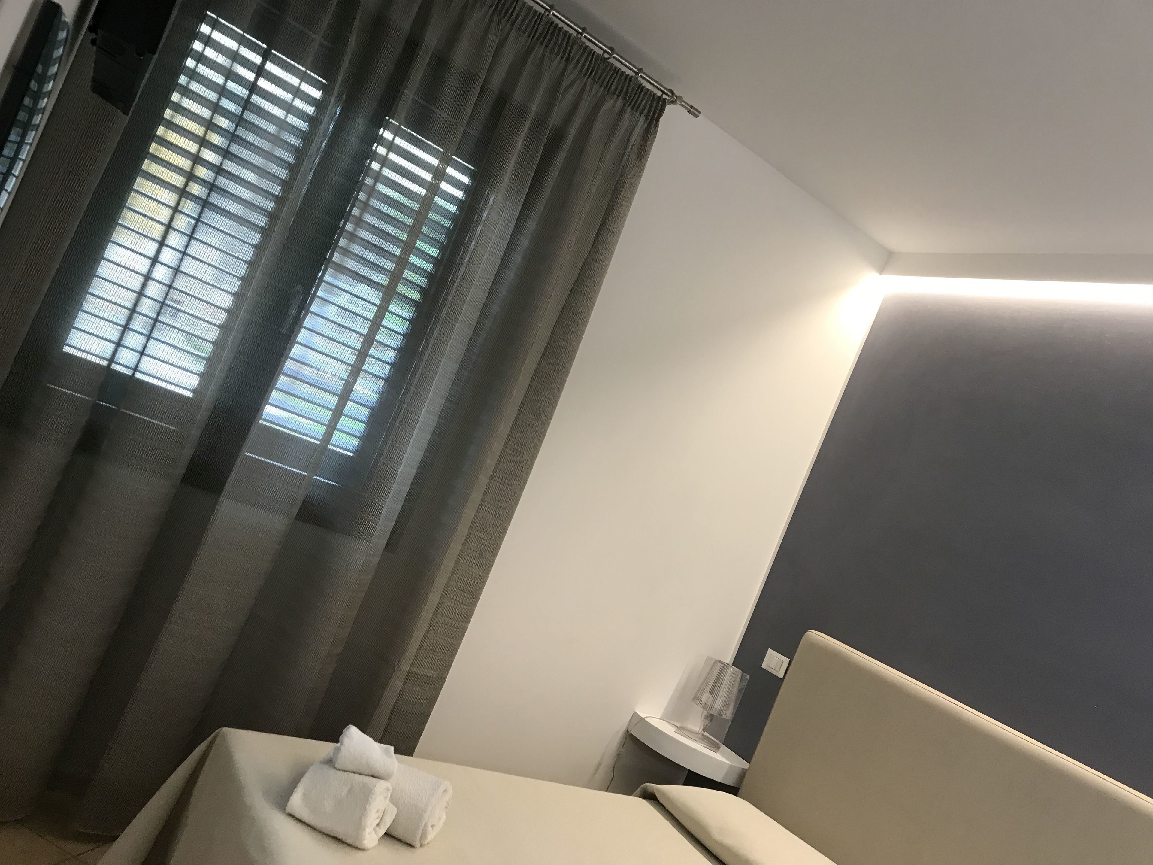 Quarto de hotel com cama, mesa de cabeceira, luminária e janela com cortinas.
