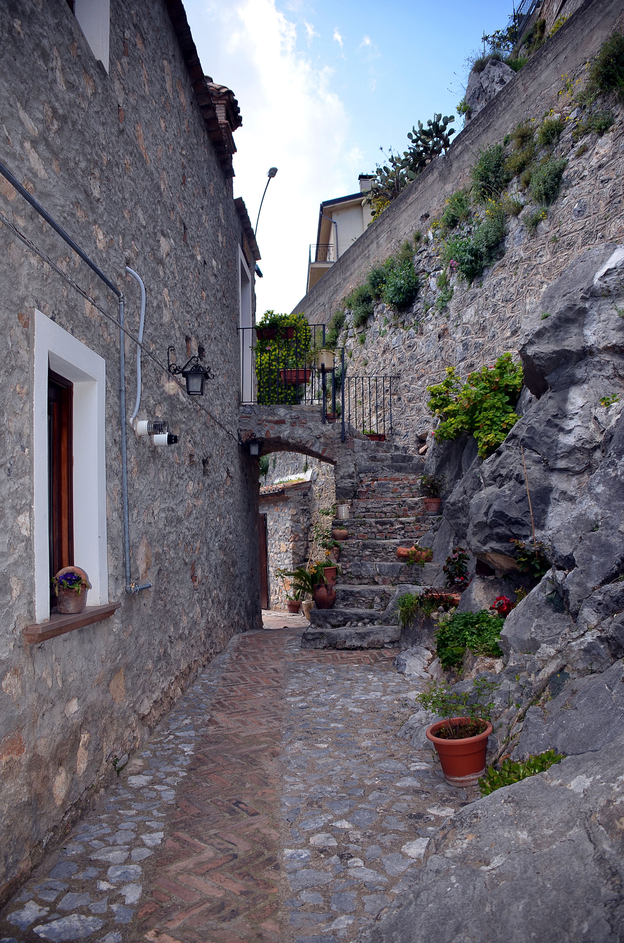 Rua de paralelepípedos estreita, ladeada por edifícios de pedra com varandas e plantas em vasos.