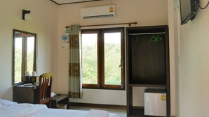 Quarto de hotel com cama, escrivaninha com cadeira, armário, janela com cortinas, aparelho de ar condicionado e televisão fixada na parede.