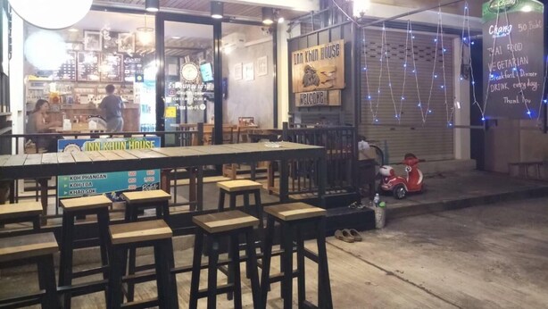 Uma área de estar externa com mesas e bancos de madeira, uma placa para Inn Khun House e um letreiro de neon anunciando comida tailandesa.