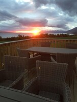 Um deck com mobiliário de vime com vista para um pôr do sol sobre um lago e montanhas.