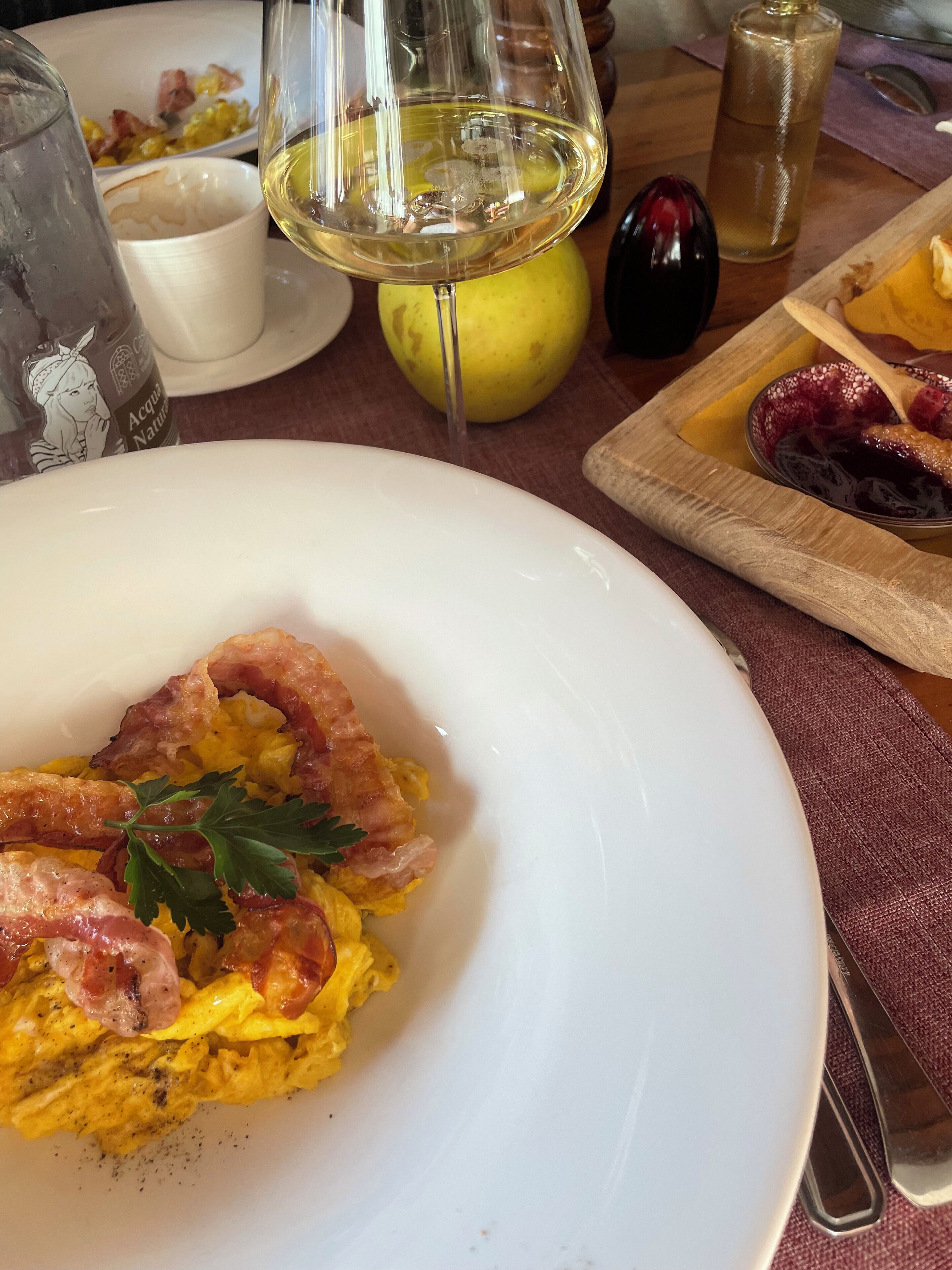 Uma mesa de café da manhã com ovos mexidos cobertos com bacon e salsa, um copo de vinho branco e uma tábua de frutas.