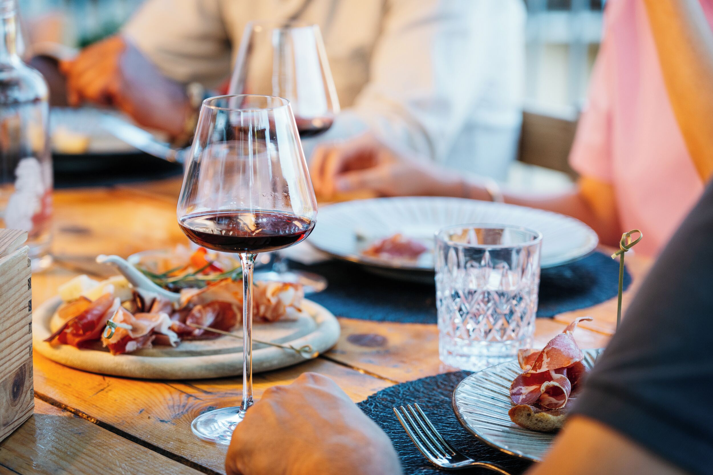 Uma mesa de madeira com um copo de vinho tinto, pratos de comida e um copo transparente.