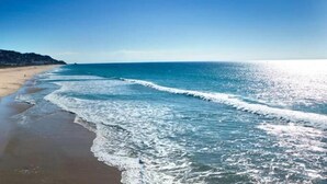 Uma extensa praia de areia com ondas suaves, céu claro e uma colina ao longe.