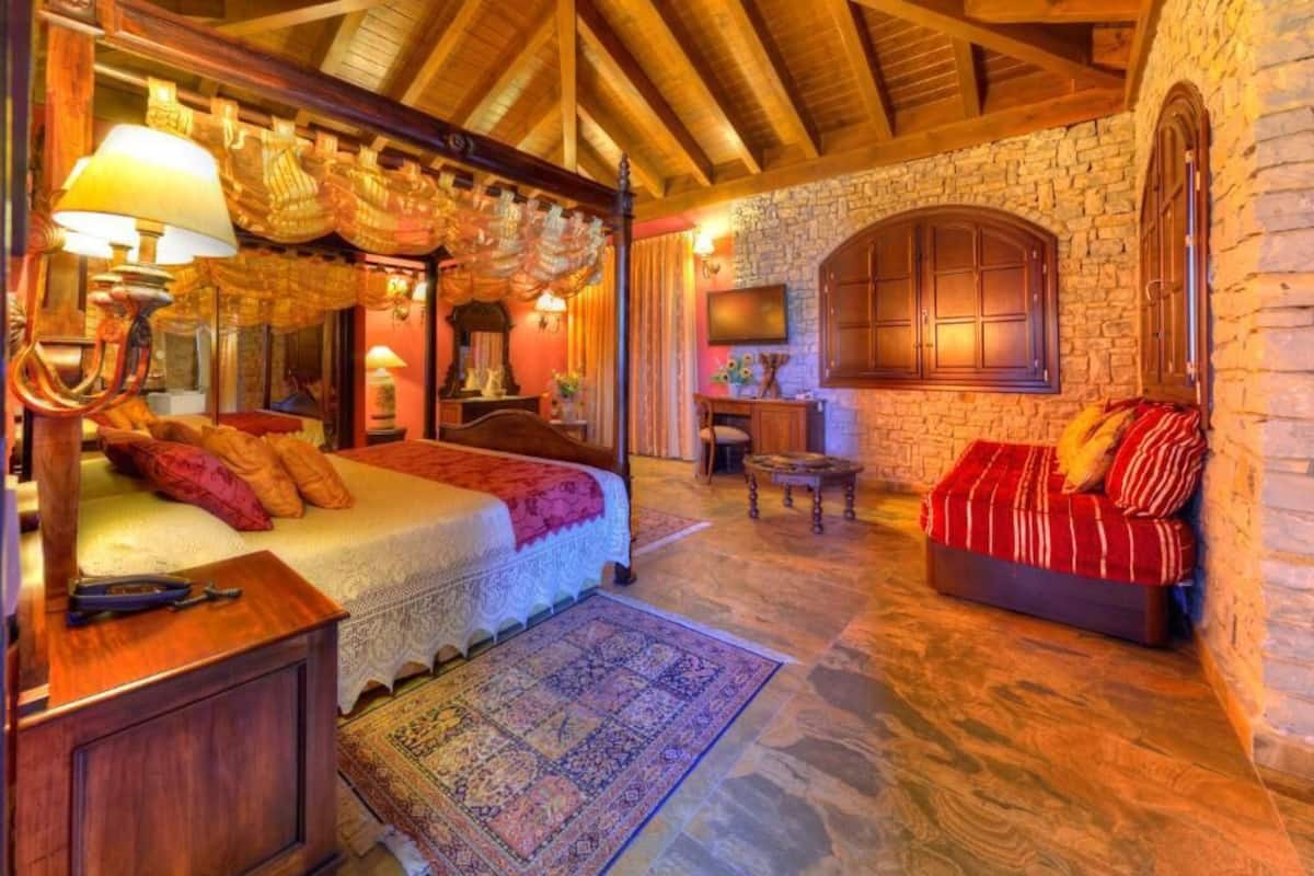 Quarto tradicional com parede de pedra, cama de madeira com roupa de cama vermelha, banco de madeira com almofadas vermelhas, uma pequena mesa e uma televisão presa na parede.
