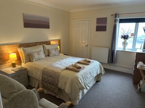 Quarto bem organizado com cabeceira de madeira, mesa de cabeceira com abajur, cama com colcha bege e almofadas, janela com vista exterior e radiador.