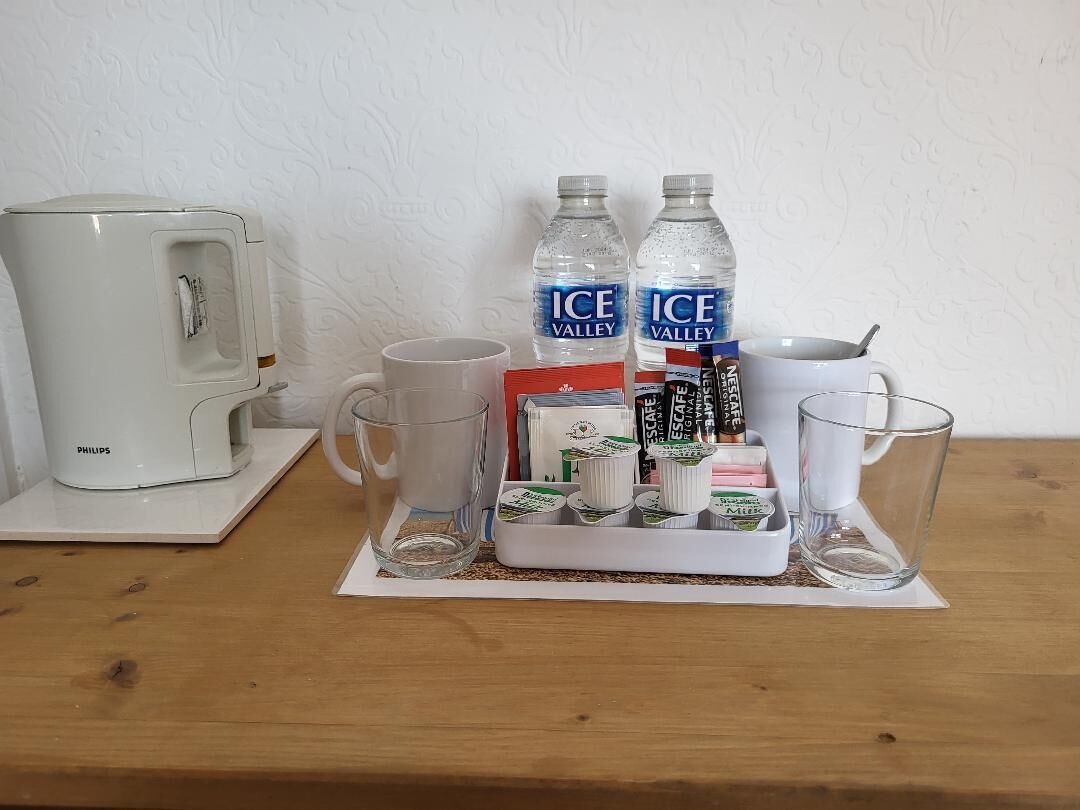 Uma cafeteira, duas garrafas de água, uma bandeja com copos e acompanhamentos, e uma caneca branca sobre uma mesa de madeira.