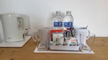 Uma cafeteira, duas garrafas de água, uma bandeja com copos e acompanhamentos, e uma caneca branca sobre uma mesa de madeira.
