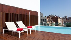 Uma área de piscina no terraço com duas espreguiçadeiras brancas e uma toalha vermelha, com vista para a paisagem urbana.
