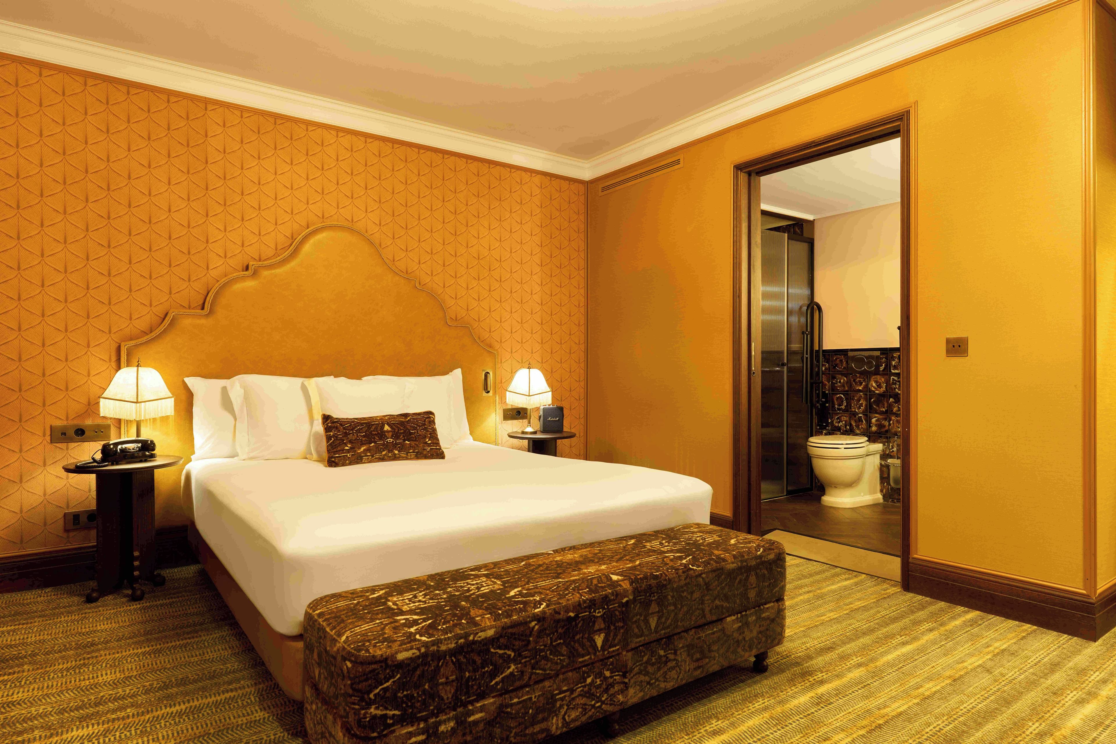 Quarto de hotel com uma cama grande, luminárias laterais, um banco de madeira, um espelho e um banheiro visível por uma porta aberta.