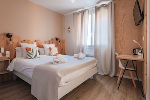 Quarto de hotel com cabeceira de madeira, cama com roupa de cama branca, escrivaninha com cadeira e janela com cortinas.
