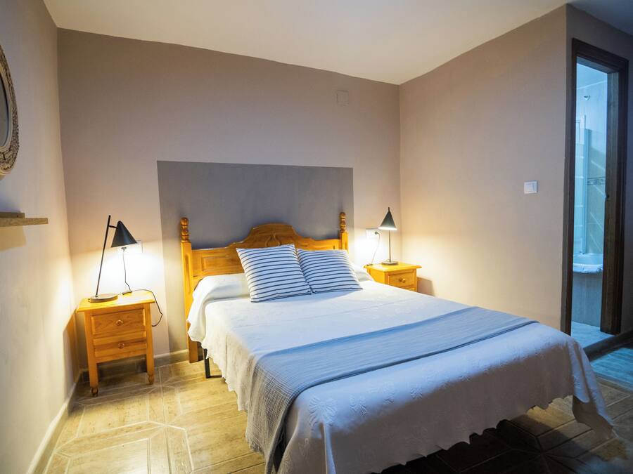 Quarto com uma cama, duas mesas de cabeceira com luminárias, um painel de cabeceira de madeira e vista para o exterior através de uma porta.