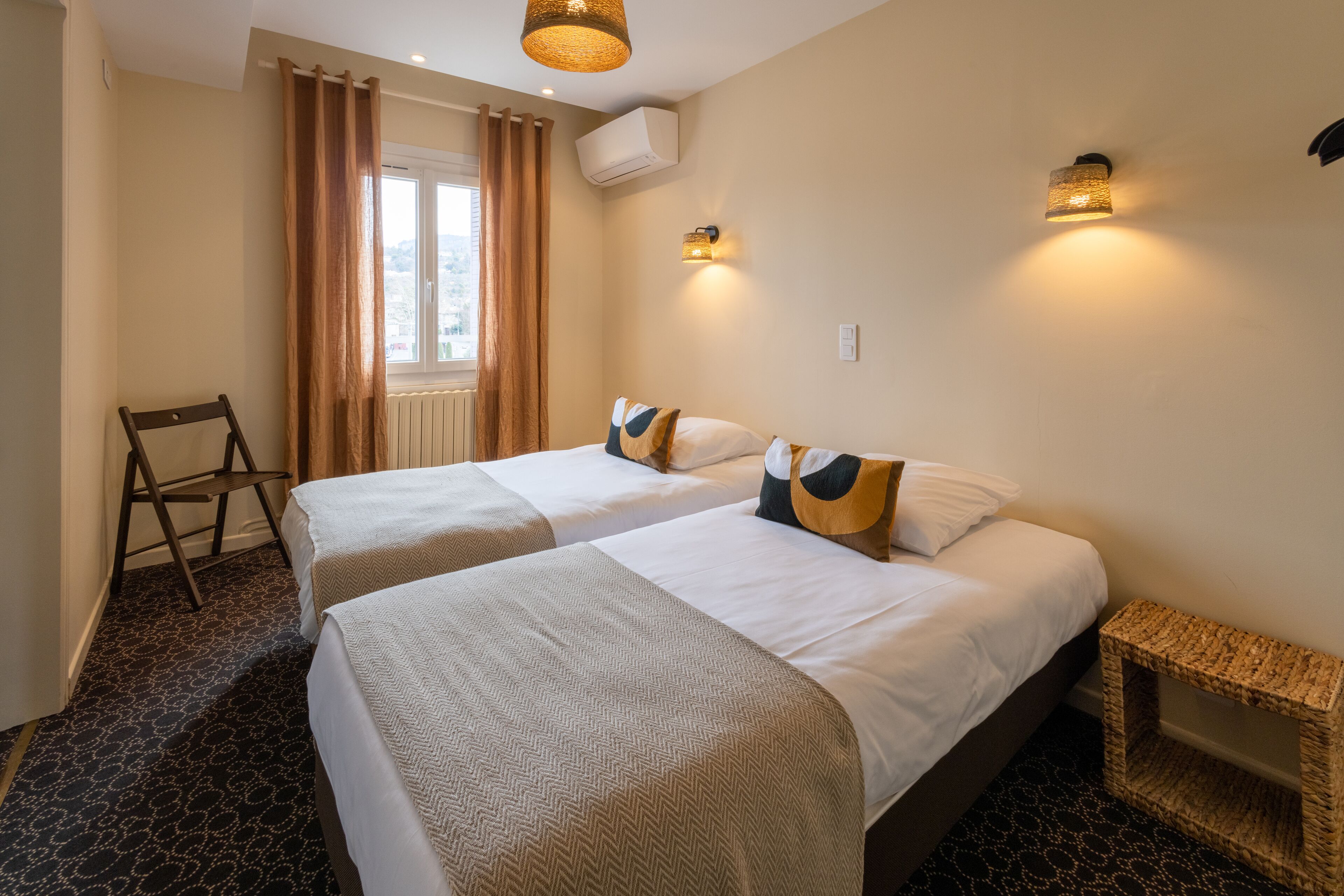Quarto de hotel com duas camas, uma mesinha, uma cadeira e uma janela com cortinas.