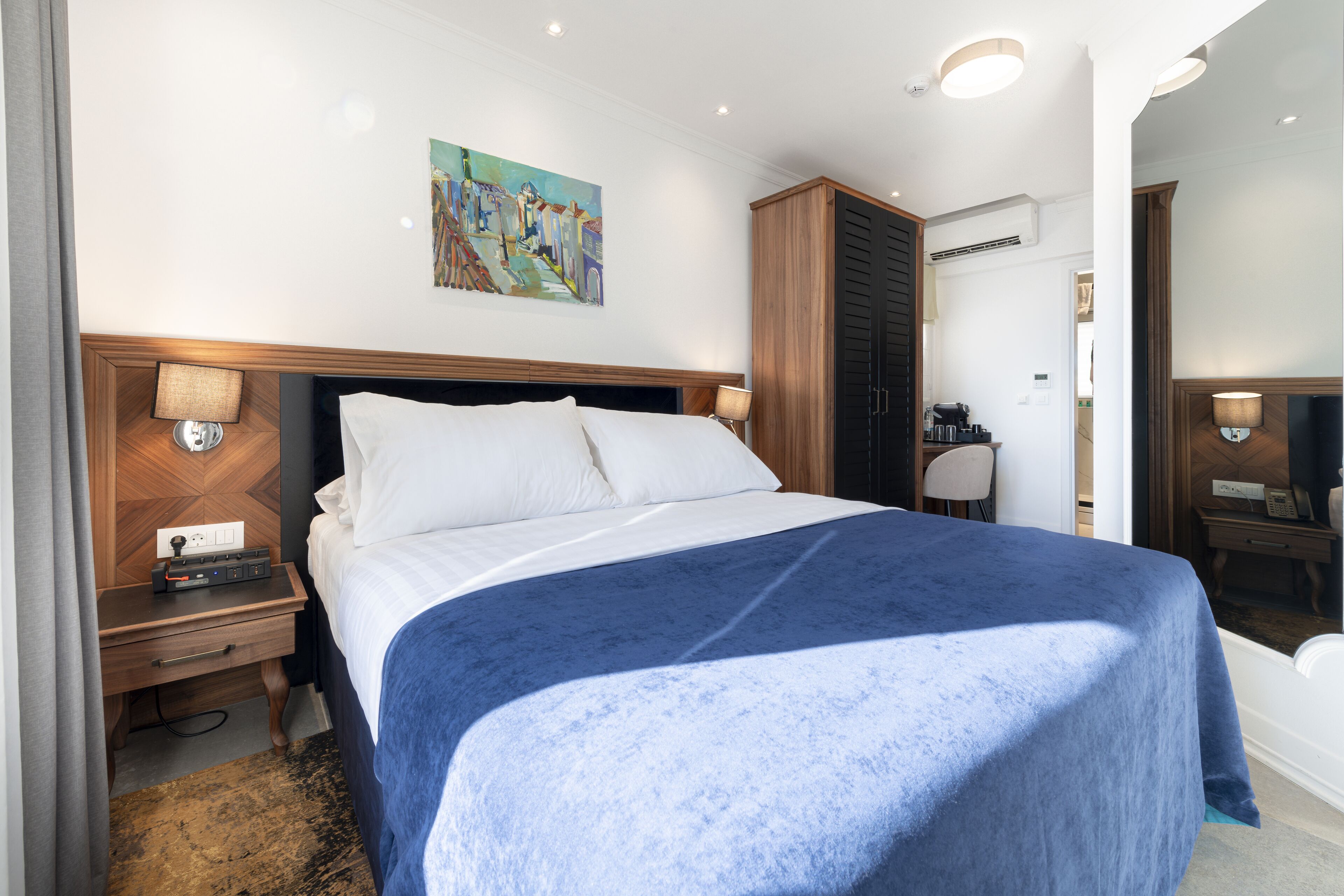 Quarto de hotel com uma cama grande, cabeceira de madeira, um quadro na parede e uma escrivaninha com luminária.