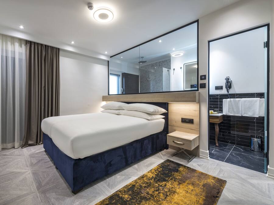 Quarto moderno com uma cama grande, mesas de cabeceira, um espelho e banheiro com chuveiro e pia.