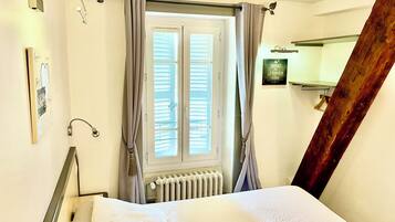 Quarto com cama, radiador, janela com persianas, luz acoplada à parede e uma viga de madeira.