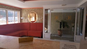 Uma área de recepção com um sofá vermelho, entrada por uma porta de vidro, uma planta em vaso e uma placa indicando serviços para hóspedes.