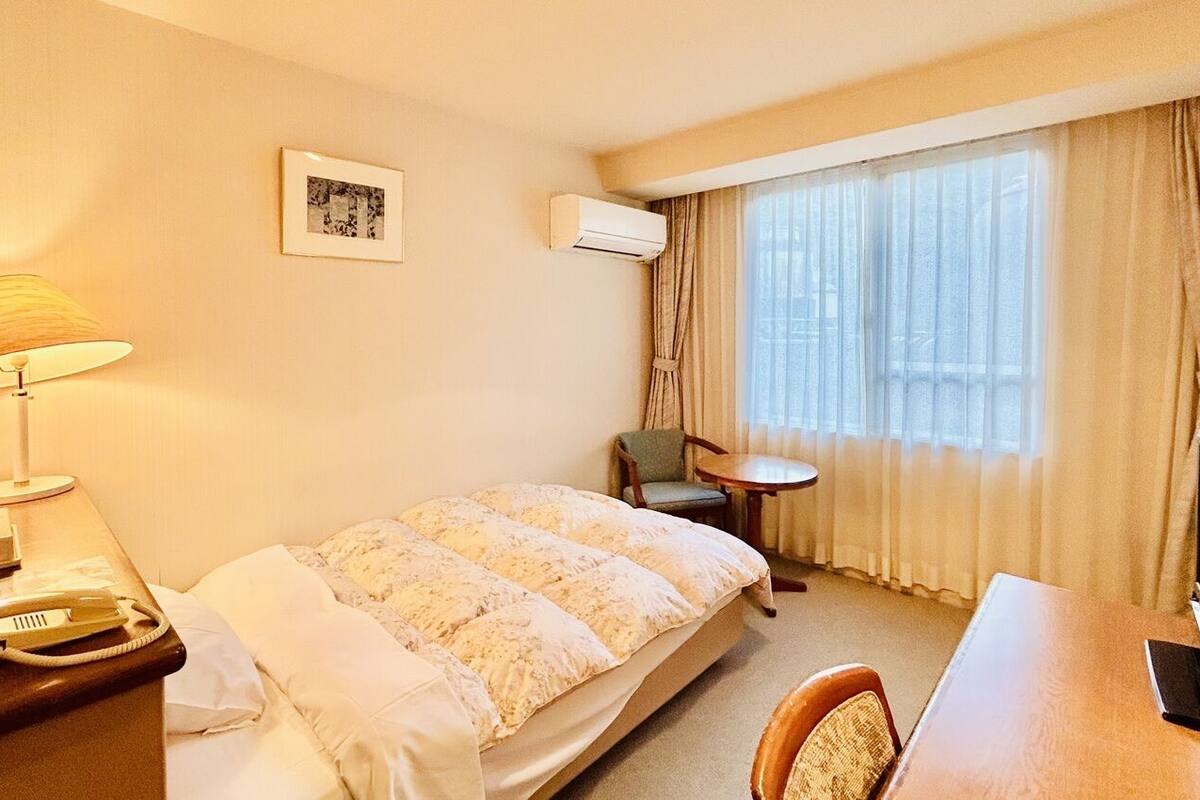 Quarto de hotel com cama, escrivaninha com computador, cadeira, luminária, ar-condicionado e janela com cortinas.