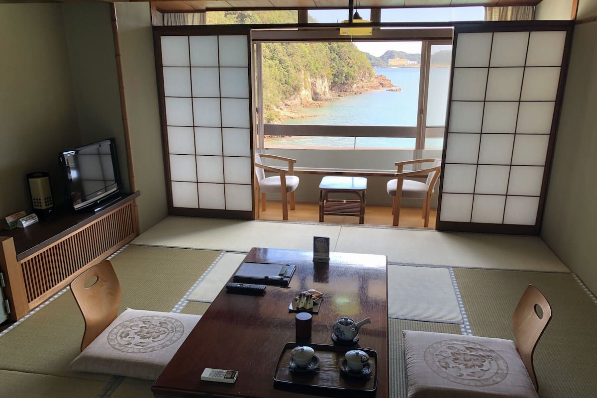 Um quarto japonês tradicional com tatames, uma mesa baixa, uma televisão e vista para a paisagem costeira.