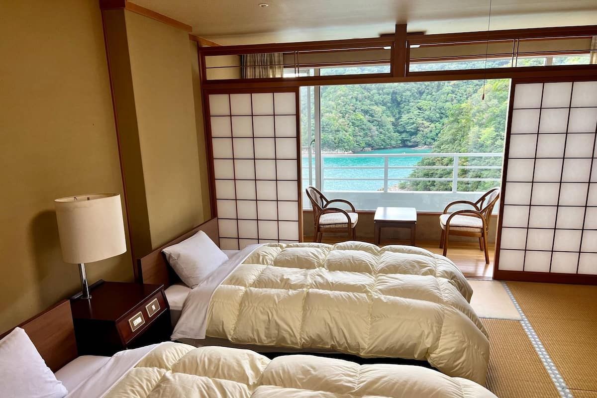 Quarto de hotel com duas camas, uma mesa de cabeceira, uma luminária, vista para o mar e uma porta japonesa tradicional deslizante.