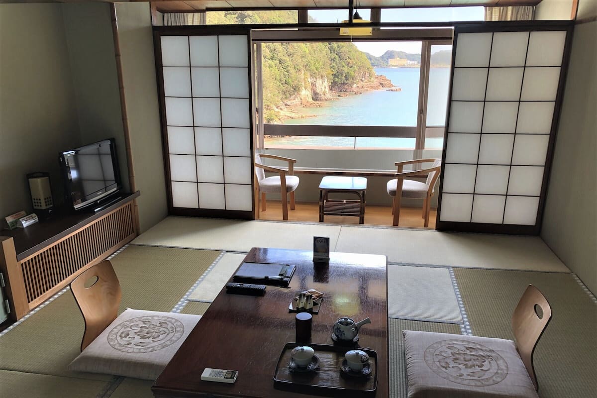 Um quarto japonês tradicional com tatames, uma mesa baixa com jogo de chá, uma televisão em um móvel e vista para a paisagem costeira.