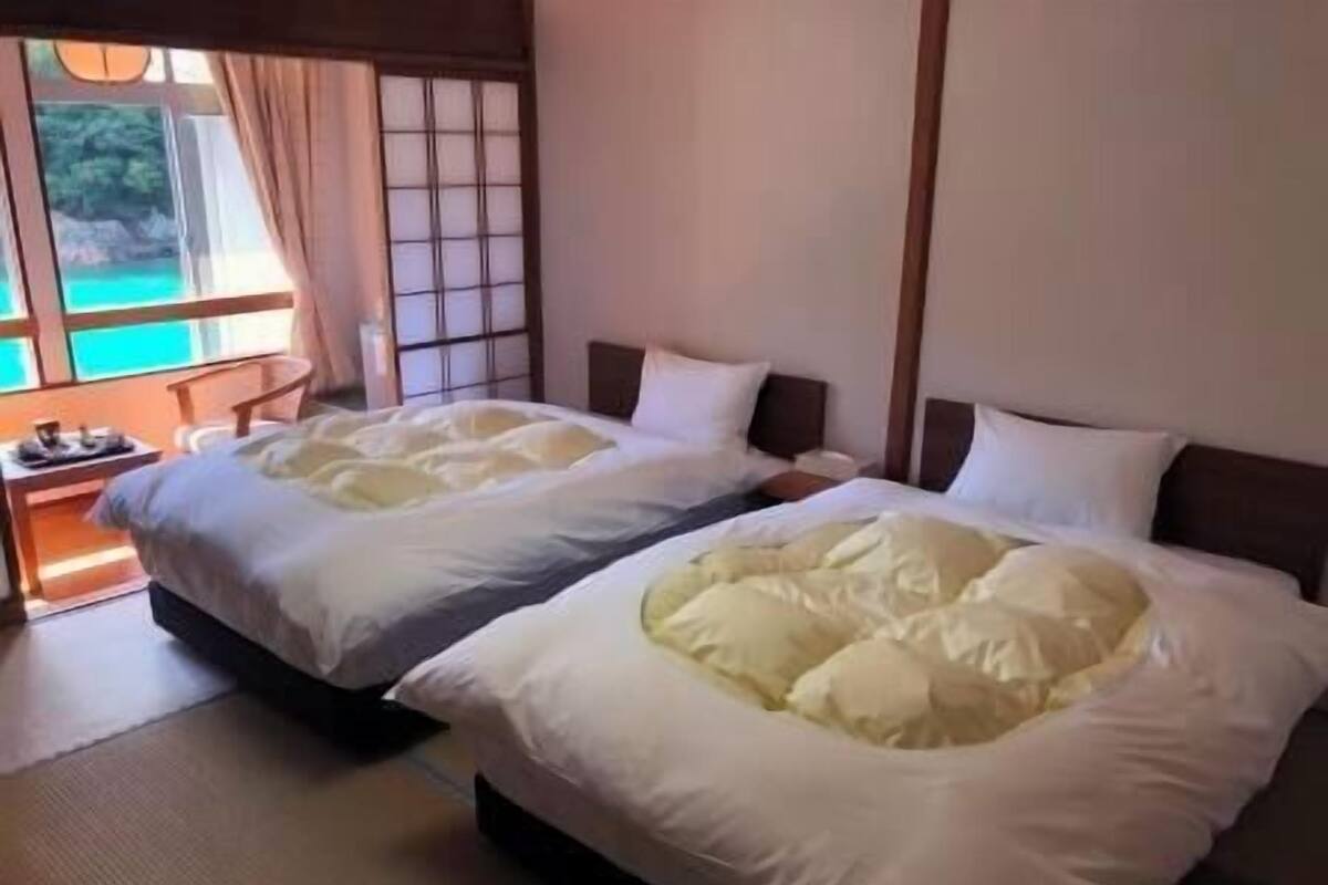 Quarto de hotel com duas camas, cabeceira de madeira, uma mesinha com abajur e vista para uma paisagem natural pela janela.