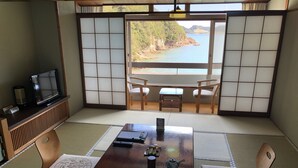 Um quarto japonês tradicional com tatames, uma mesa baixa, uma televisão e vista para a paisagem costeira.