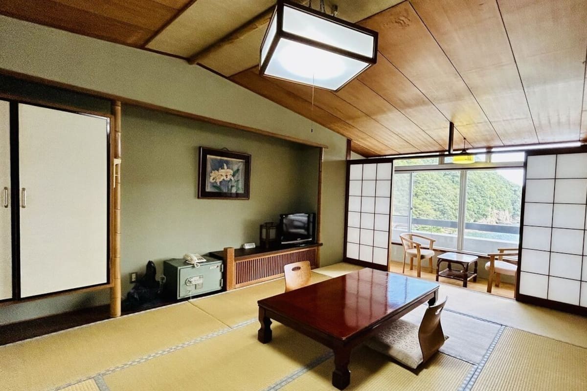 Um quarto japonês tradicional com tatames, uma mesa de madeira, portas deslizantes, uma televisão e vista para o verde lá fora.