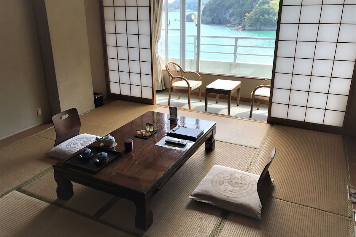 Um quarto japonês tradicional com piso de tatame, uma mesa de madeira baixa e vista para a paisagem costeira através de biombos shoji.