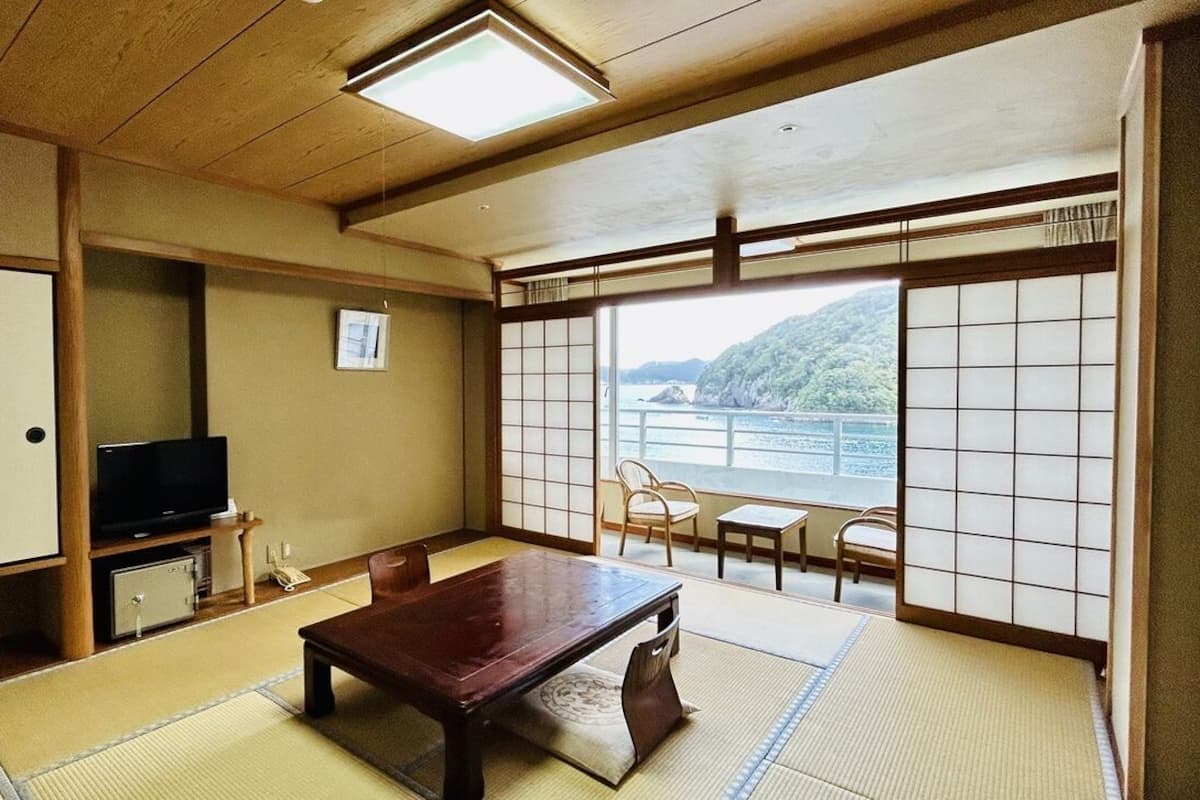 Um quarto japonês tradicional com tatames, uma mesa de madeira, uma televisão e portas deslizantes que dão para uma varanda com vista para um corpo de água e colinas.