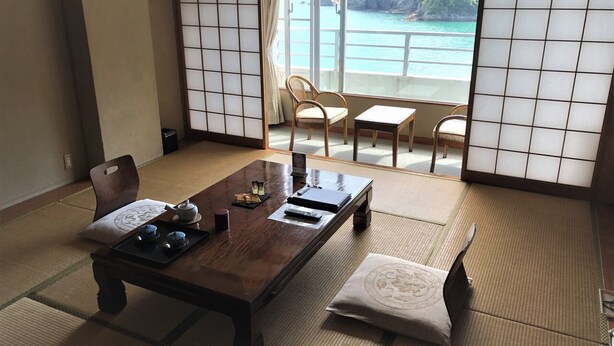 Um quarto japonês tradicional com piso de tatame, uma mesa de madeira baixa e vista para a paisagem costeira através de biombos shoji.
