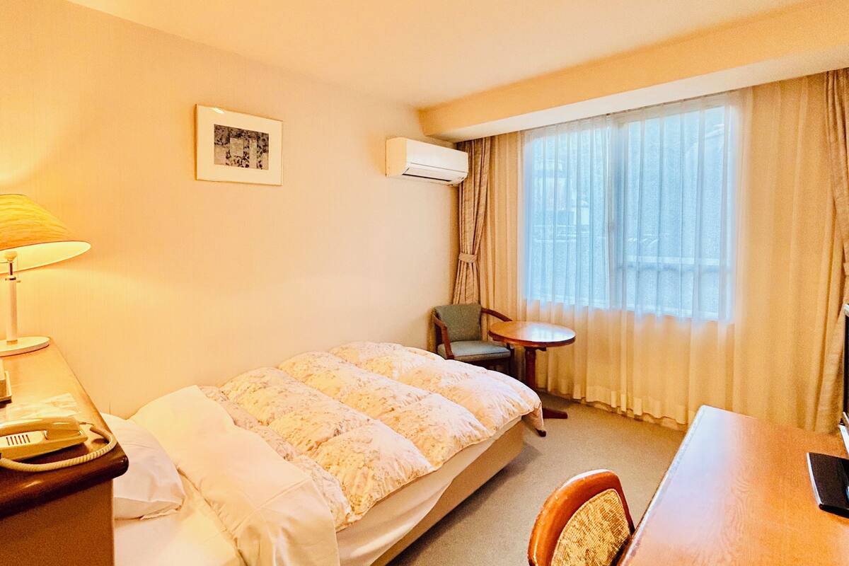 Quarto de hotel com cama, escrivaninha, cadeira, luminária, ar condicionado e janela com cortinas.