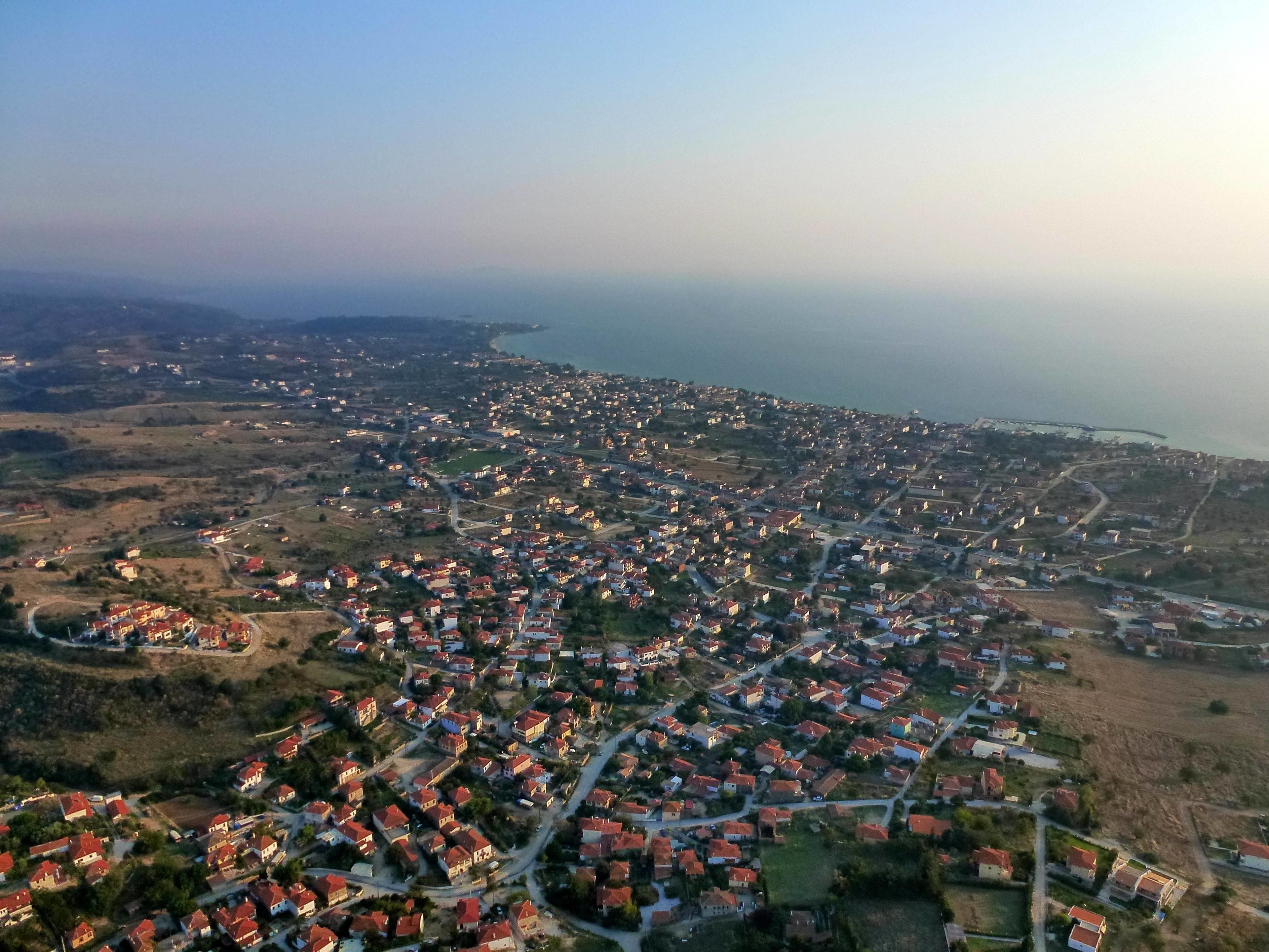 Vista aérea de uma vila costeira com um conjunto denso de edifícios com telhados vermelhos, uma linha costeira visível e uma mistura de áreas residenciais e espaços abertos.