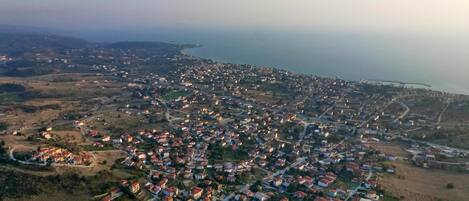 Vista aérea de uma vila costeira com um conjunto denso de edifícios com telhados vermelhos, uma linha costeira visível e uma mistura de áreas residenciais e espaços abertos.