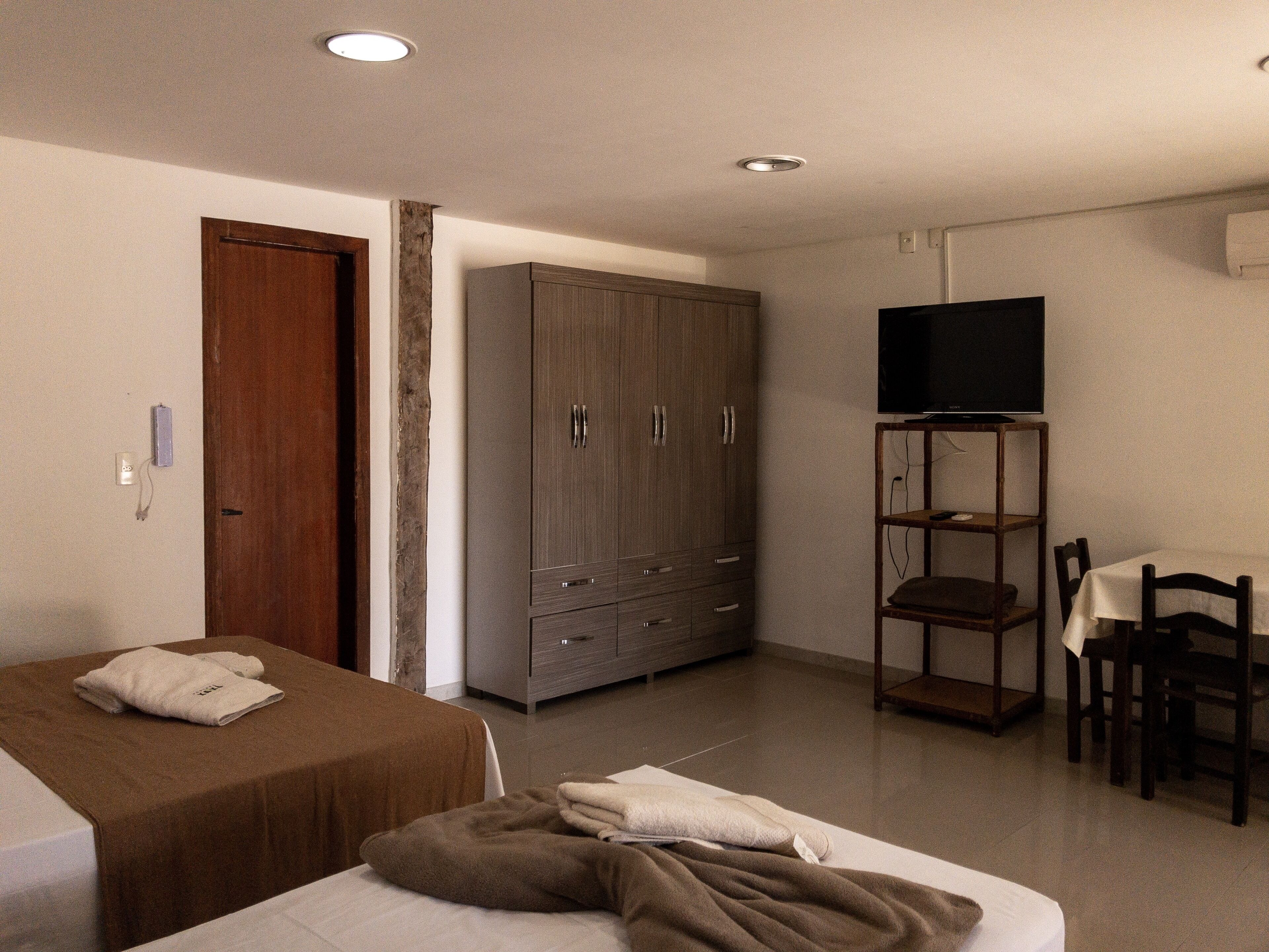 Quarto de hotel com duas camas, um guarda-roupa, uma televisão sobre um suporte e uma pequena mesa com cadeiras.