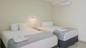 Quarto de hotel com duas camas, cada uma com roupa de cama branca e saia cinza. Possui um ar condicionado fixado na parede e uma pequena mesa com uma garrafa e um copo.