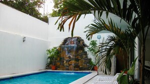 Piscina com cachoeira de pedra, cercada por paredes brancas e plantas em vasos.