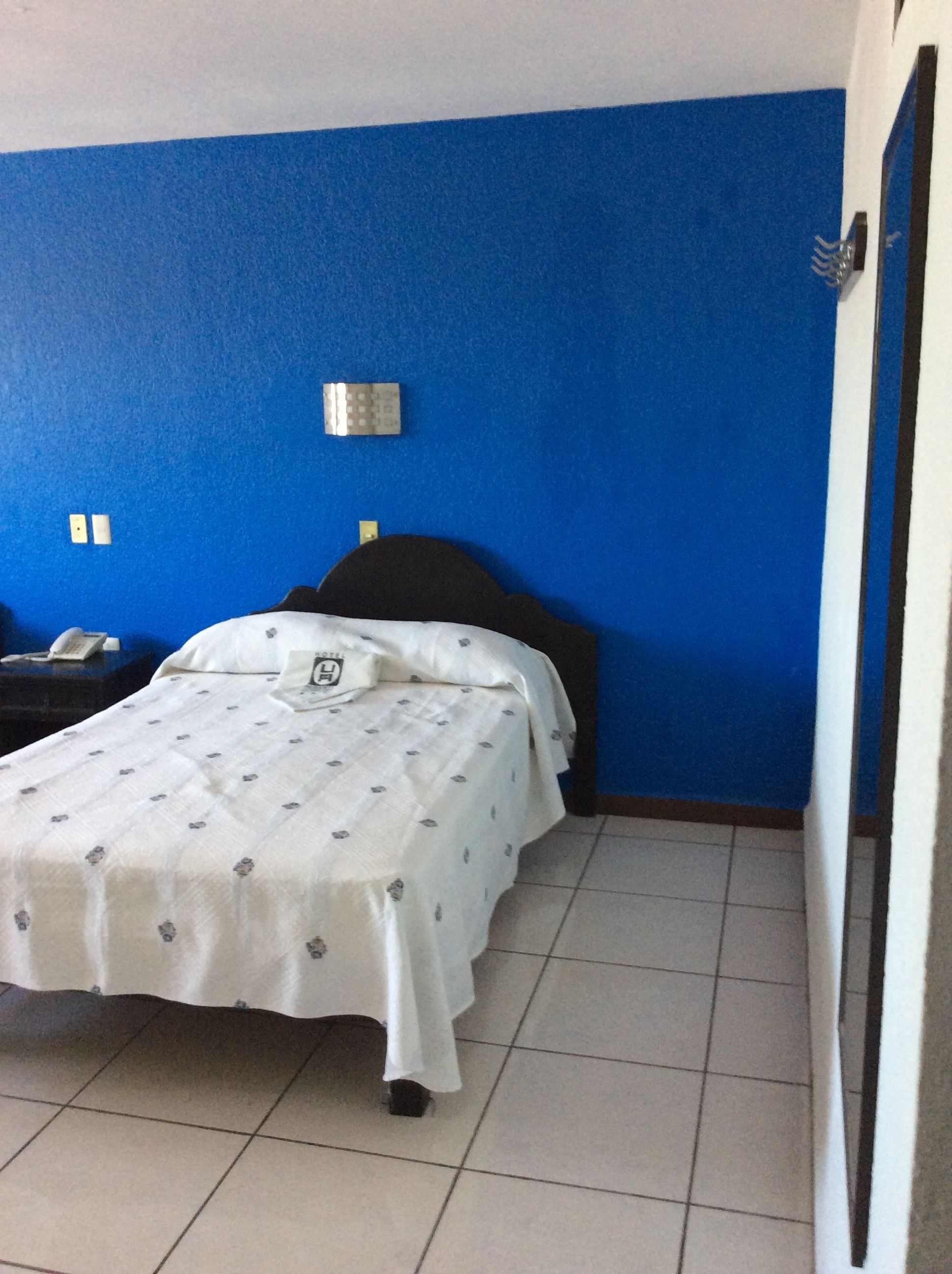 Quarto de hotel com parede azul, cama com colcha branca, cabeceira preta e uma pequena mesa de cabeceira com telefone.