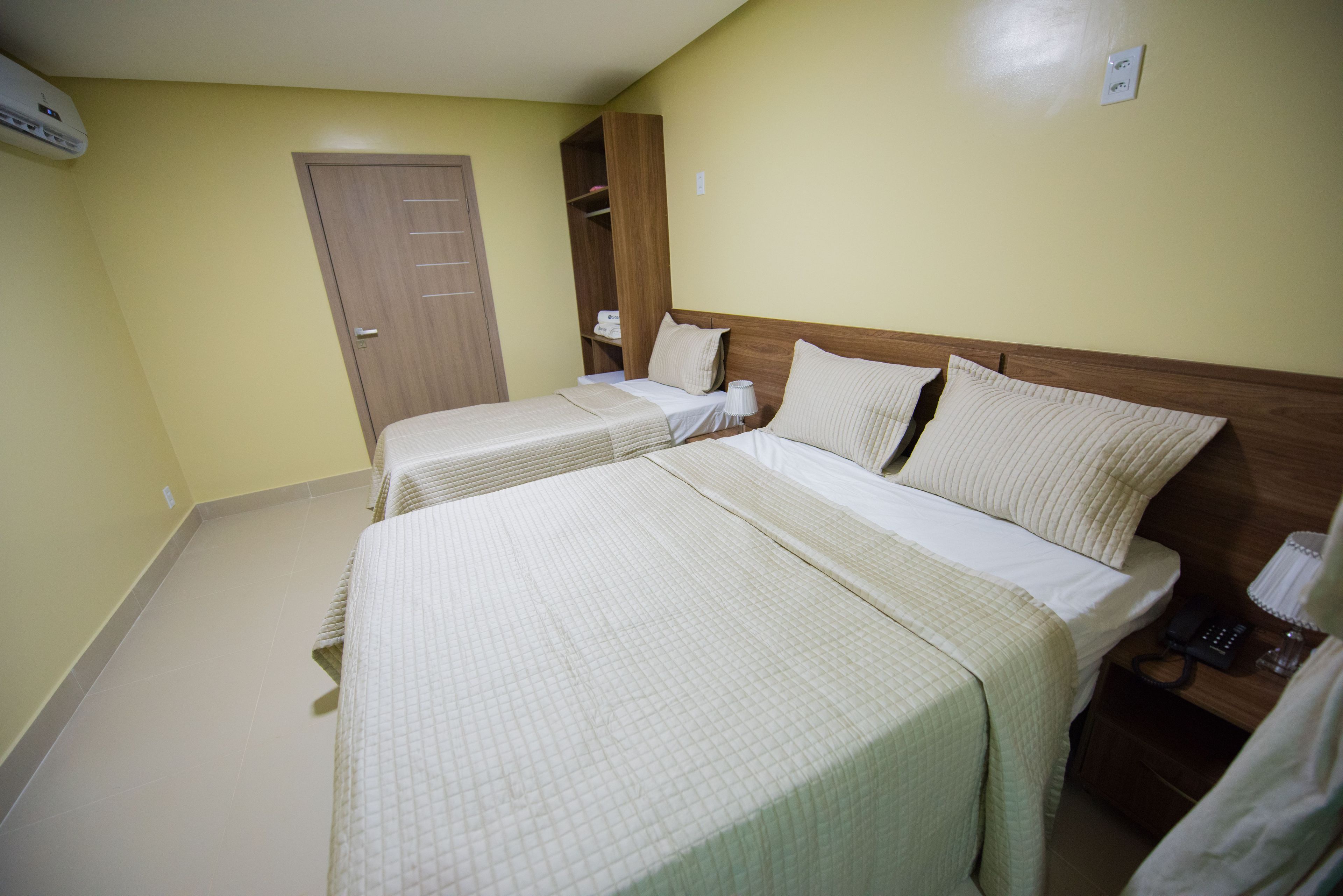 Quarto de hotel com duas camas, um guarda-roupa de madeira, uma mesa de cabeceira com abajur e um telefone.
