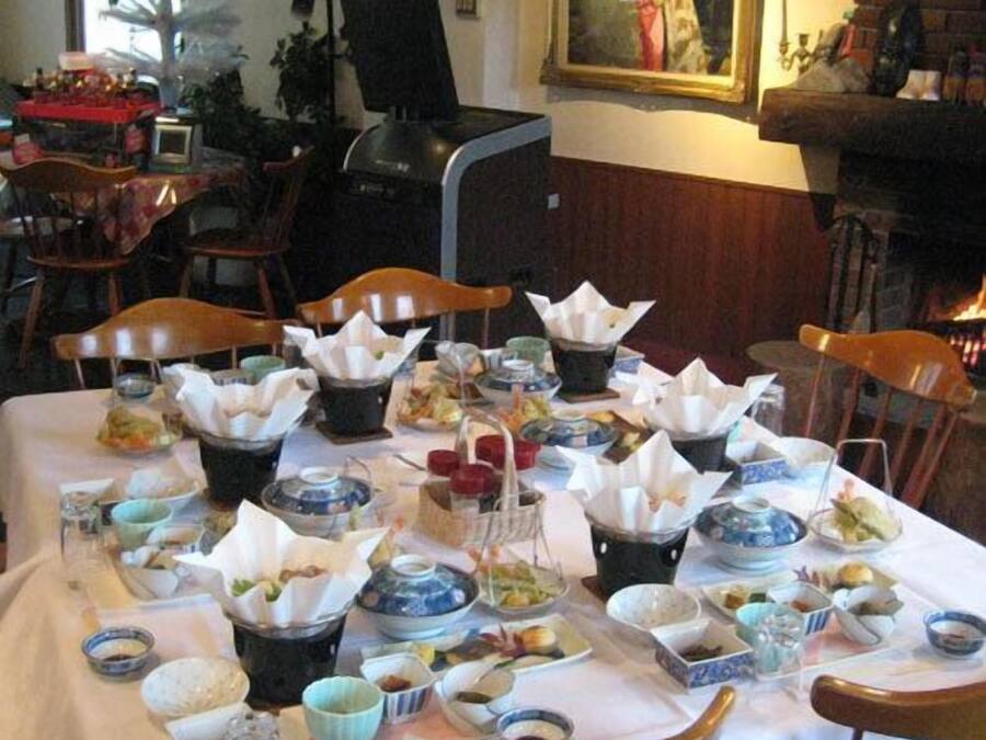 Uma sala de jantar com várias mesas preparadas para refeição, cadeiras ao redor e uma lareira ao fundo.