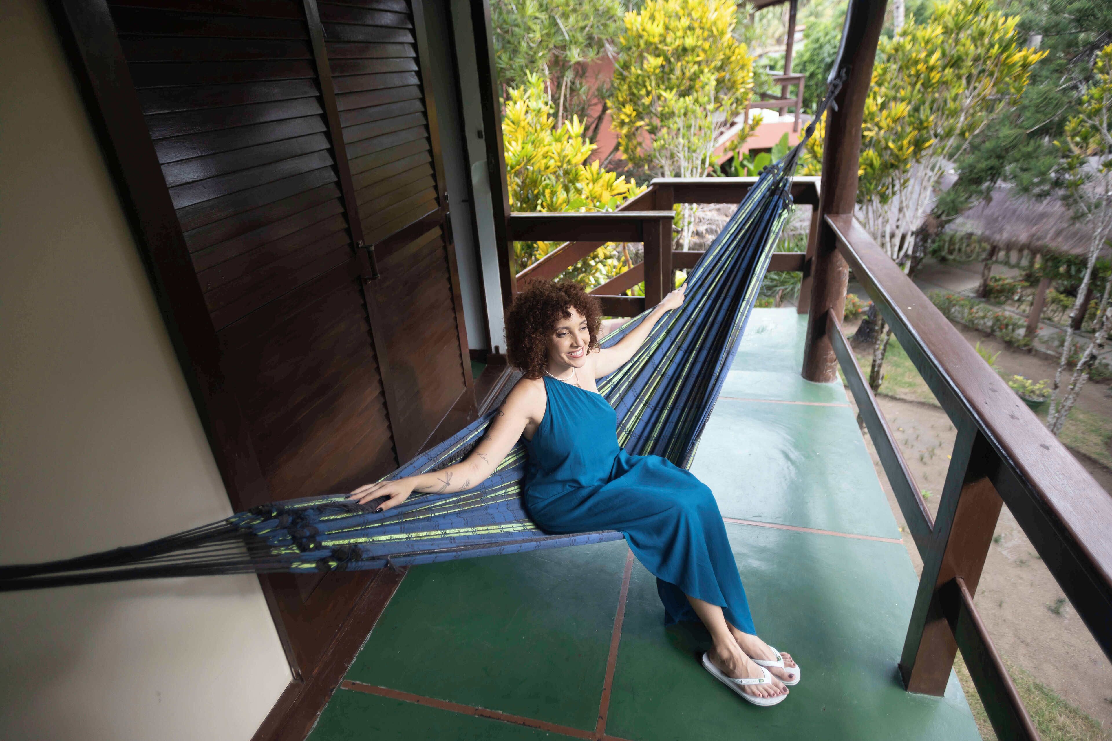 Uma pessoa relaxando em uma rede em um alpendre, com vista para vegetação e uma construção de telhado de palha ao fundo.