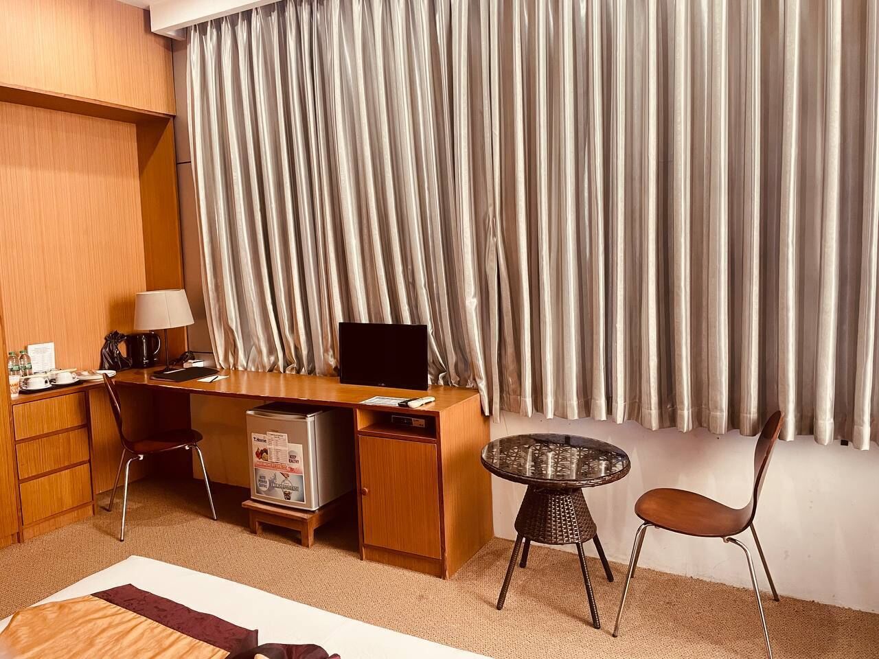 Quarto de hotel com escrivaninha de madeira, televisão, pequeno frigorífico, máquina de café, abajur, mesa redonda e duas cadeiras.