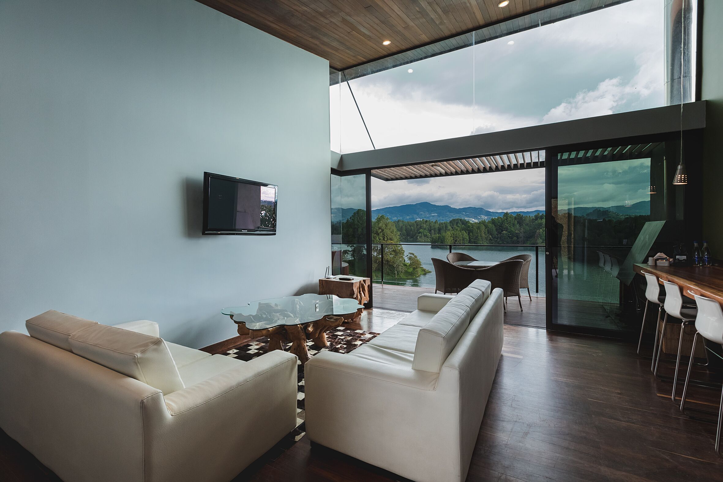Uma sala de estar moderna com parede de vidro, televisão de tela plana e área de jantar com mesa e cadeiras de madeira.