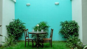 Pequena área de pátio fechada com uma mesa redonda e duas cadeiras, cercada por plantas verdes exuberantes e uma parede azul-turquesa.