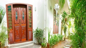 Uma porta de madeira tradicional com esculturas detalhadas e painéis de vidro colorido, ladeada por vegetação exuberante e um caminho de pedra que leva a um edifício branco.