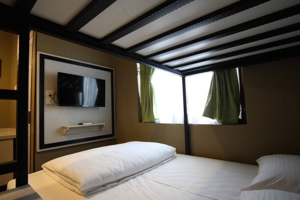 Quarto de hotel com cama, televisão presa na parede e janela com cortinas.