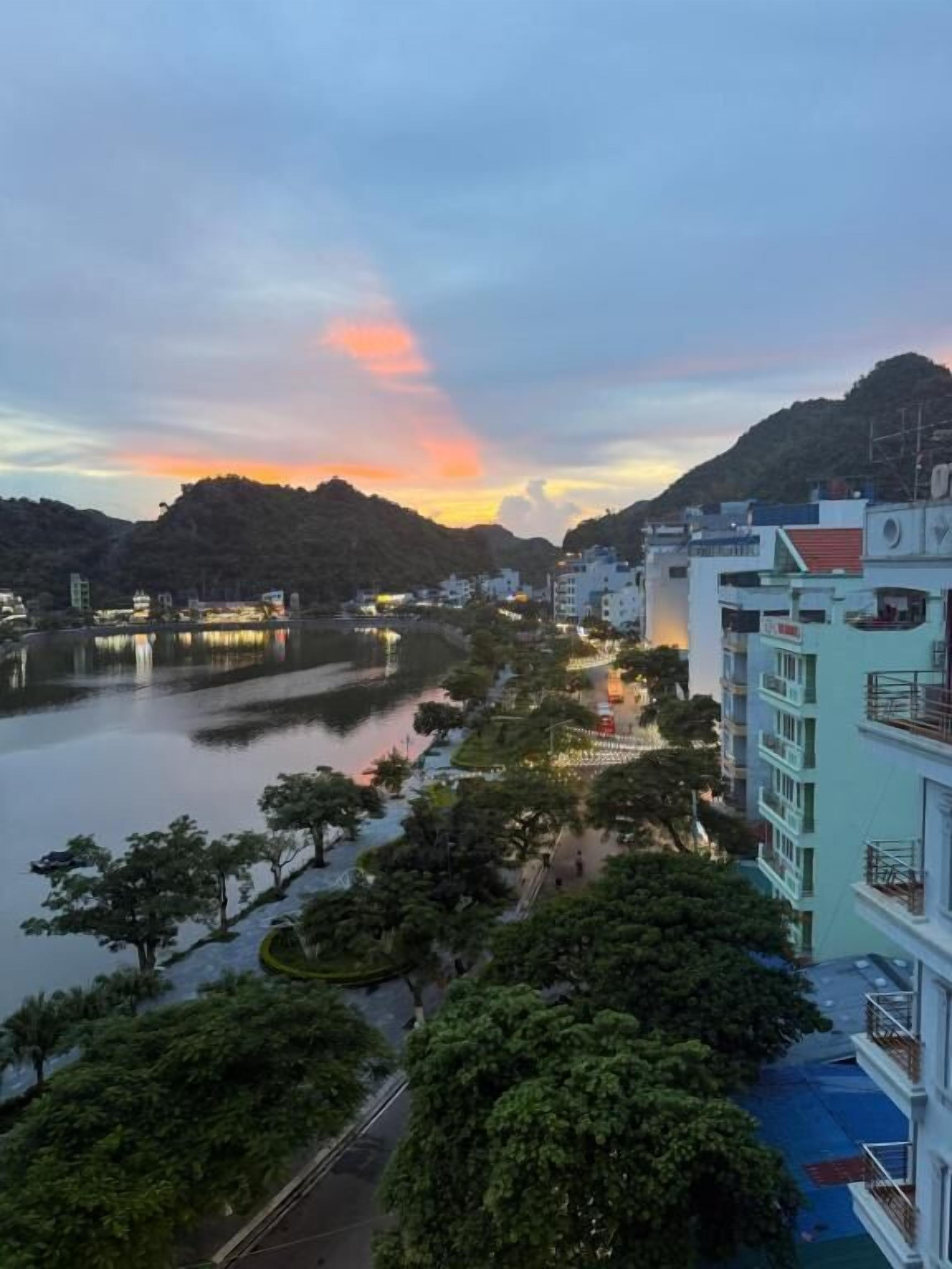 Vista ribeirinha com edifícios, árvores e um pôr do sol.