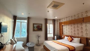 Quarto de hotel com cama, escrivaninha, cadeira, mesa pequena, ventoinha e janela com cortinas.
