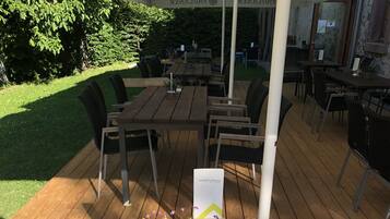 Uma área de refeições ao ar livre com mesas e cadeiras de madeira, uma variedade de comidas e bebidas, e um jardim ao fundo.